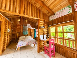 Interior de uma cabana de madeira com cama, escrivaninha, cadeira e grandes janelas.