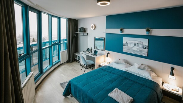 Quarto de hotel moderno com uma cama grande, uma mesa e um computador. Há uma janela com cortinas, uma luminária fixada na parede e uma moldura com uma imagem.