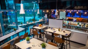Um restaurante moderno com paredes de vidro, várias mesas com cadeiras, plantas em vasos e um espaço de bar com prateleiras cheias de garrafas e copos.