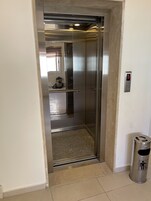 Um elevador metálico com um visor digital mostrando o número 3, corrimão e uma lixeira com símbolo de reciclagem.
