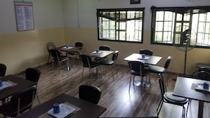 Uma sala de aula com piso de madeira, mesas com cadeiras e amplas janelas com grades.