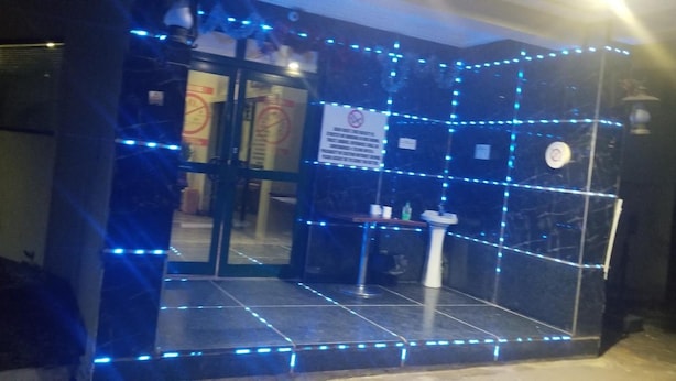 Entrada de hotel com porta de vidro, luzes LED azuis, placa de proibido fumar e um pequeno balcão de recepção.