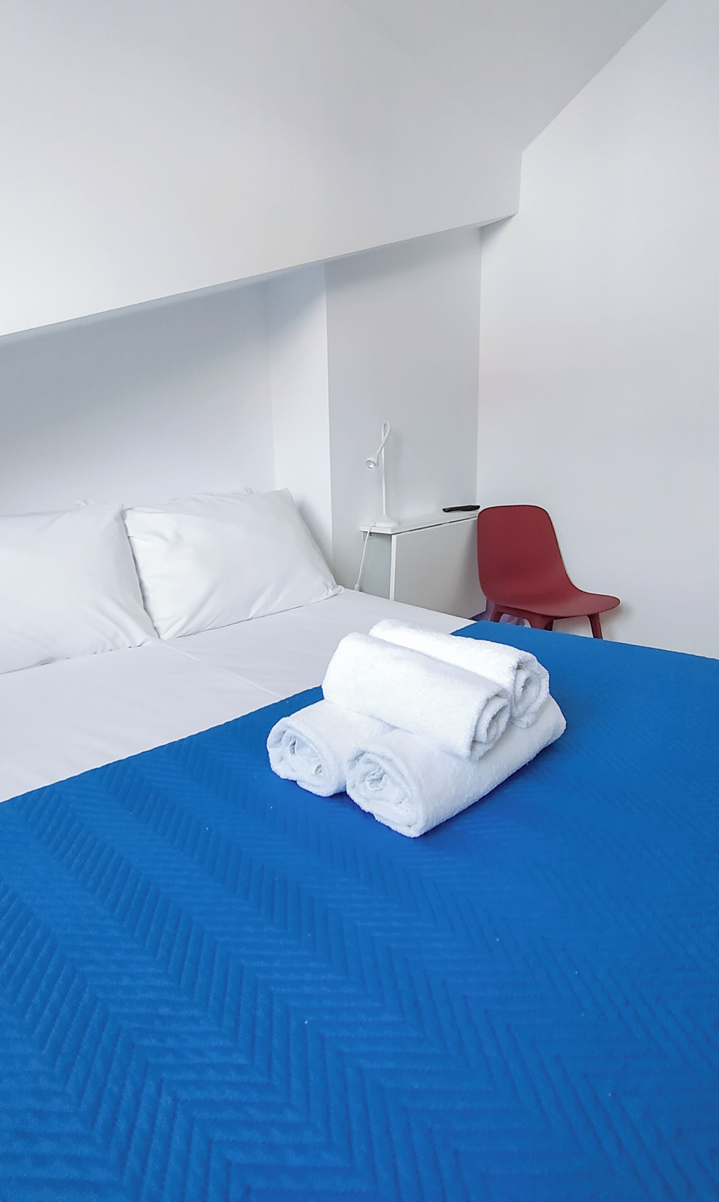 Cama bem arrumada com roupa de cama branca e colcha azul, uma cadeira vermelha e uma mesa de cabeceira branca com uma luminária.