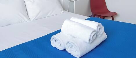 Cama bem arrumada com roupa de cama branca e colcha azul, uma cadeira vermelha e uma mesa de cabeceira branca com uma luminária.