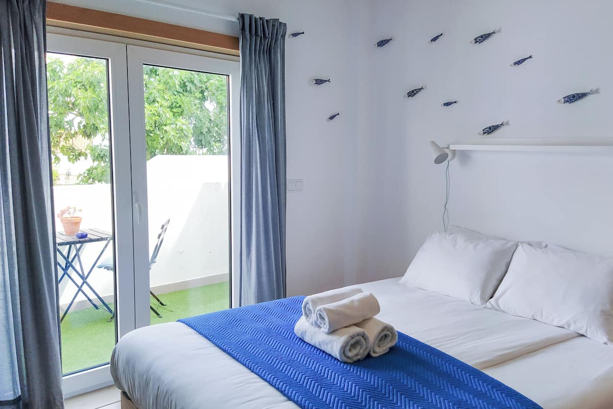 Quarto com cama, manta azul, almofadas brancas e vista para o exterior através de uma porta de correr de vidro.