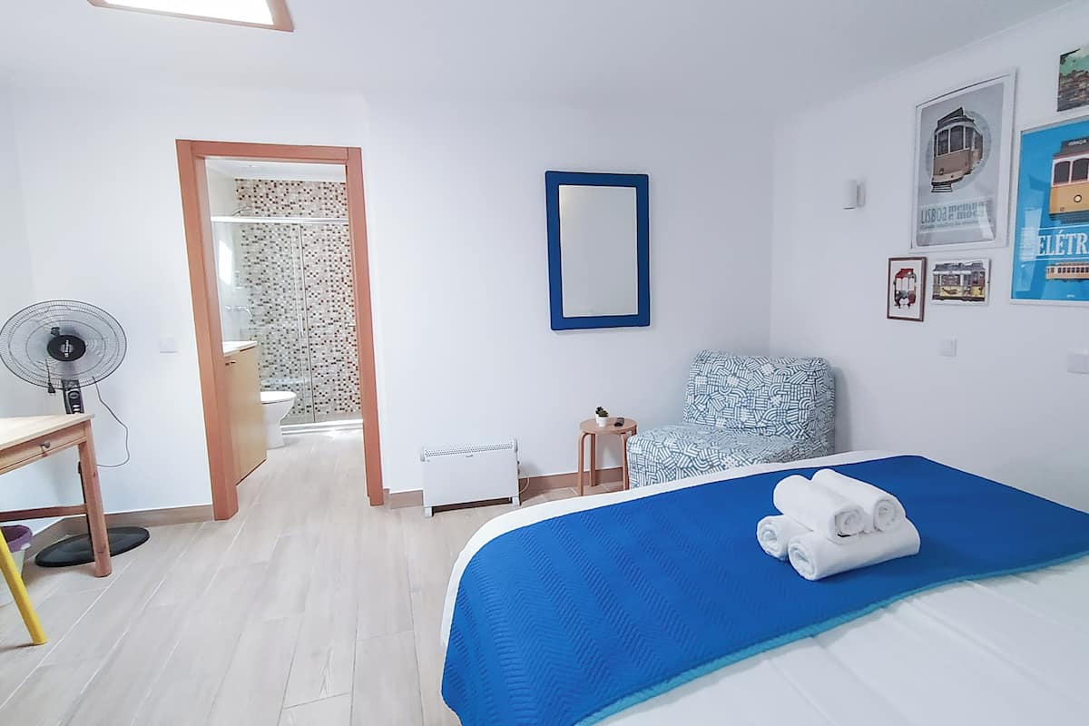 Quarto com cama, cadeira, ventoinha e uma mesinha.
