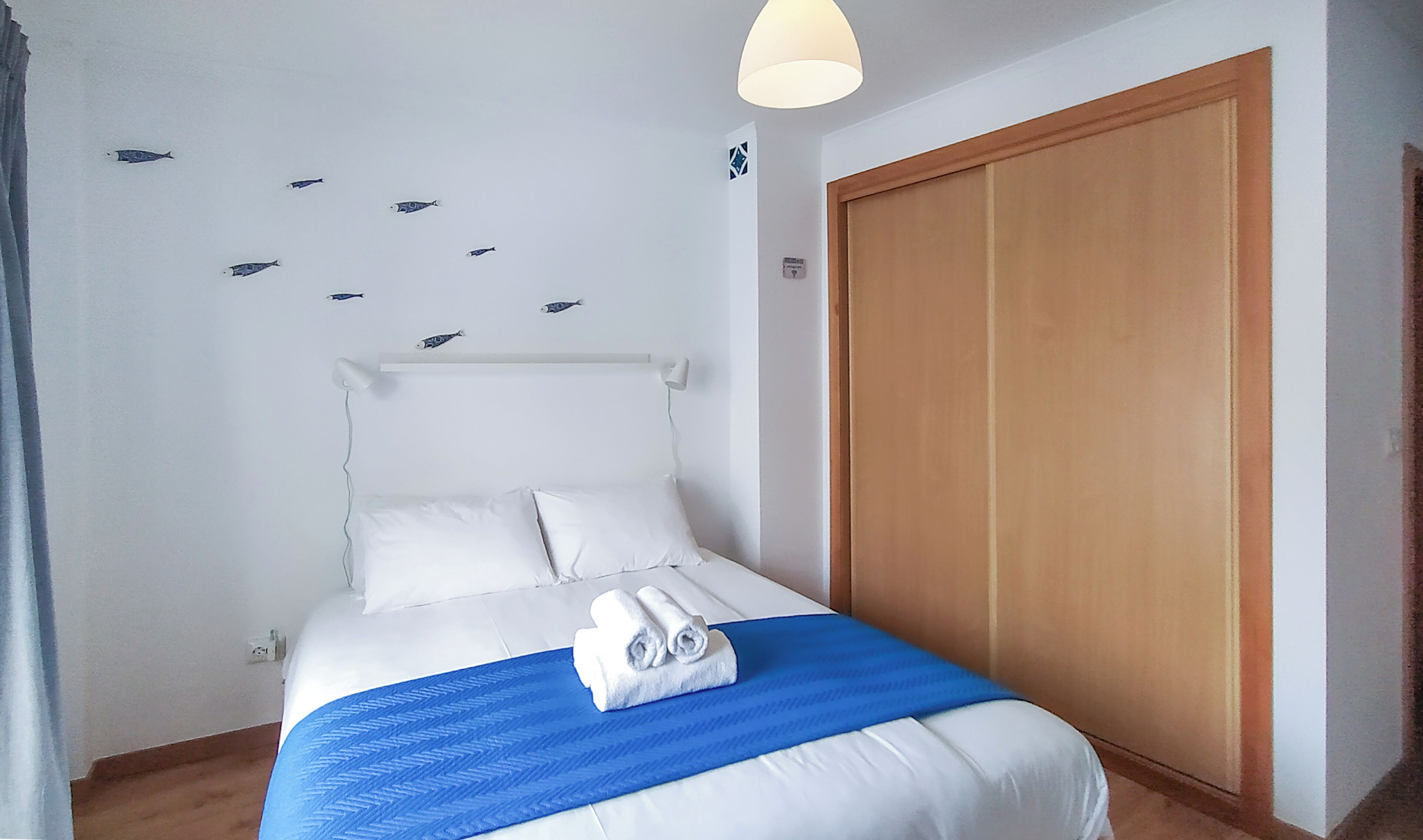 Quarto de hotel com cama, roupa de cama branca, manta azul e guarda-roupa de madeira.