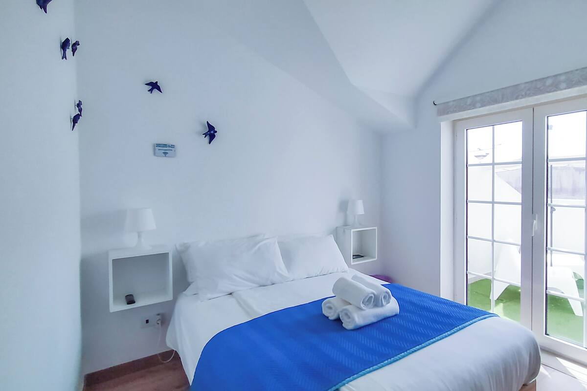 Quarto com cama, mesa de cabeceira, luminária e janela com vista para área verde lá fora.