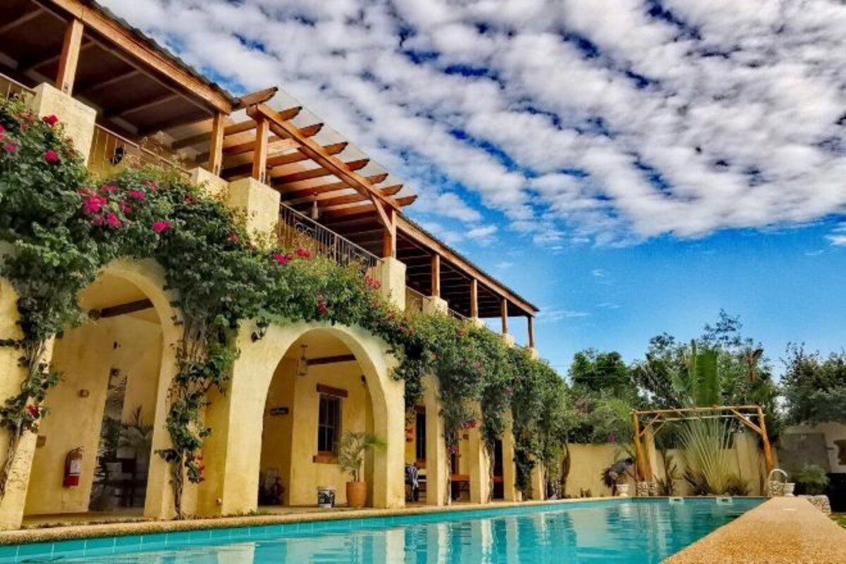 Uma área à beira da piscina com um alpendre coberto, arcos e um céu azul claro.