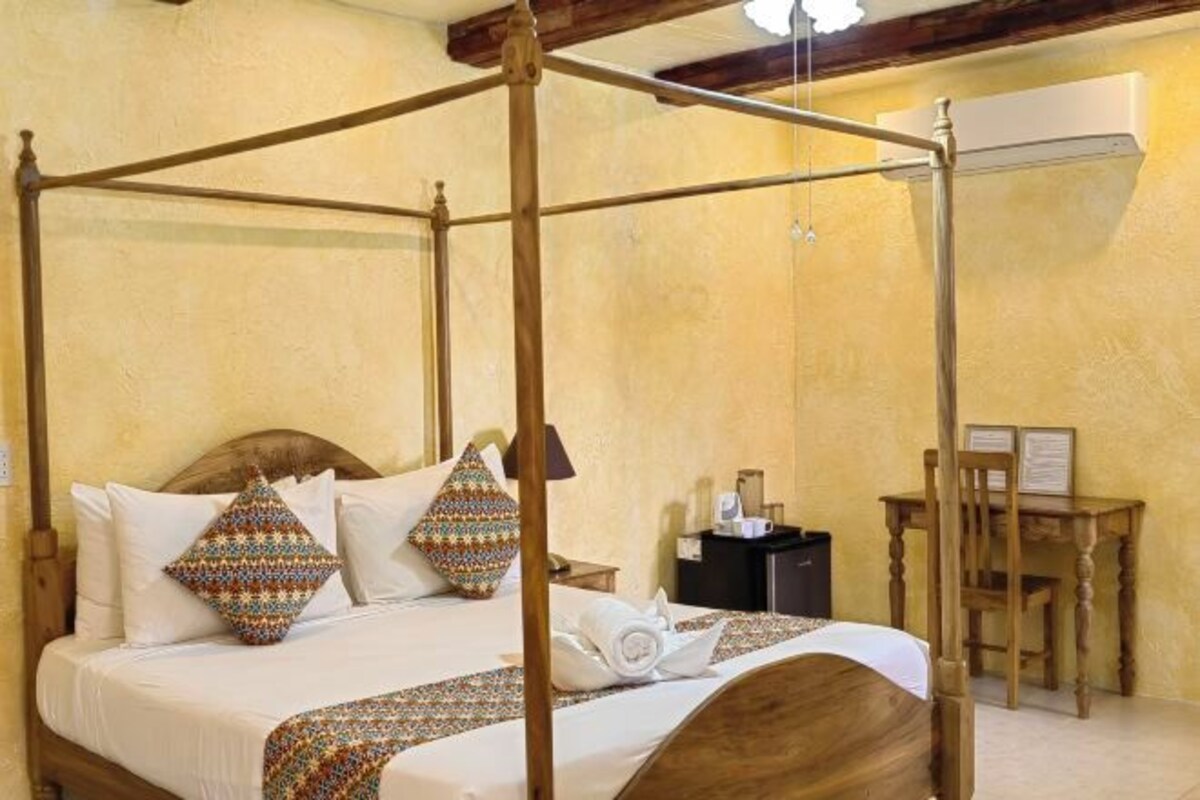 Uma cama com dossel de madeira, roupa de cama branca e almofadas estampadas. Uma pequena mesa com abajur e um certificado emoldurado fica ao lado da cama.