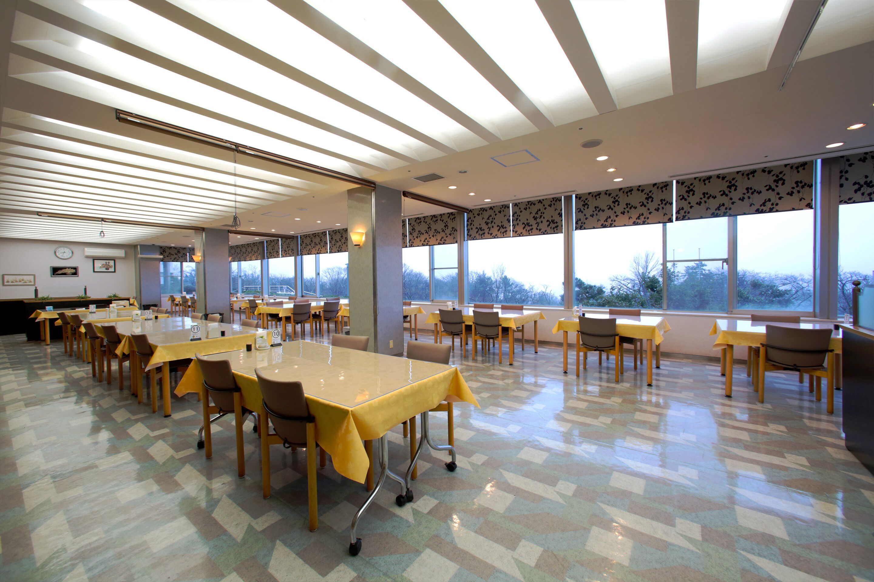 Uma sala de jantar ampla, com várias mesas e cadeiras, janelas grandes e piso com padrão.