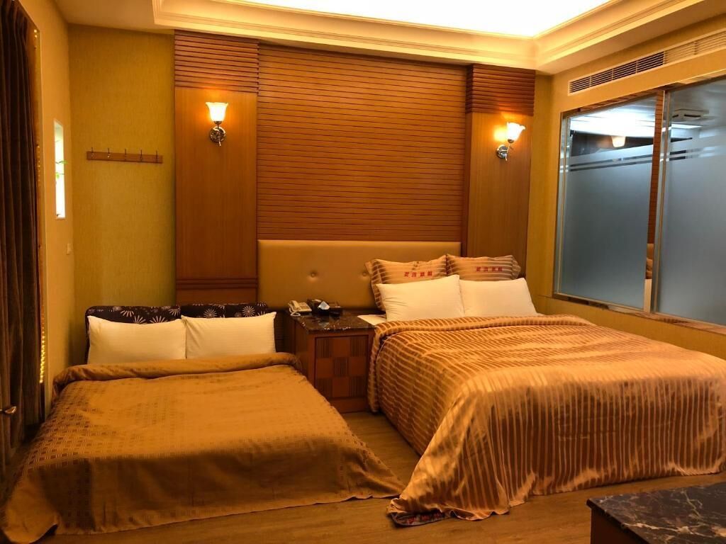 Quarto de hotel com duas camas, cabeceira de madeira, luminárias laterais, mesa de cabeceira e janela com vidro jateado.