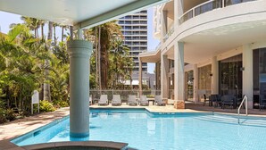 Piscina com espreguiçadeiras e hidromassagem, cercada por palmeiras e um edifício moderno.