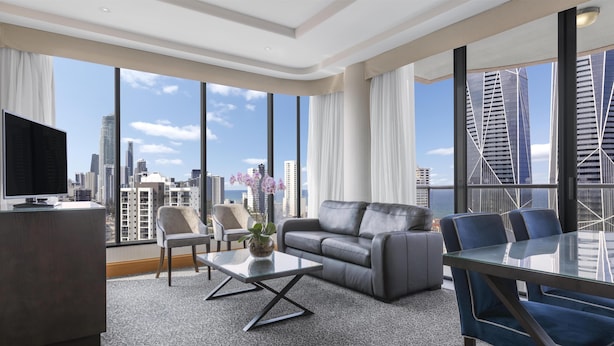 Quarto de hotel moderno com uma grande janela com vista para a cidade, um sofá, uma mesa de centro de vidro e uma TV sobre um suporte.