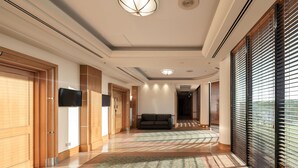 Corredor moderno de hotel com portas de madeira, televisão de tela plana, sofá e janelas amplas com persianas.