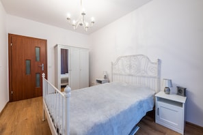 Quarto com cama branca, porta de madeira, guarda-roupa e mesa de cabeceira com abajur.