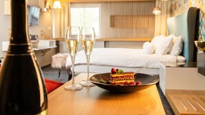 Uma mesa de jantar com um prato de bolo e duas taças de vinho, uma garrafa de vinho e uma cama ao fundo.
