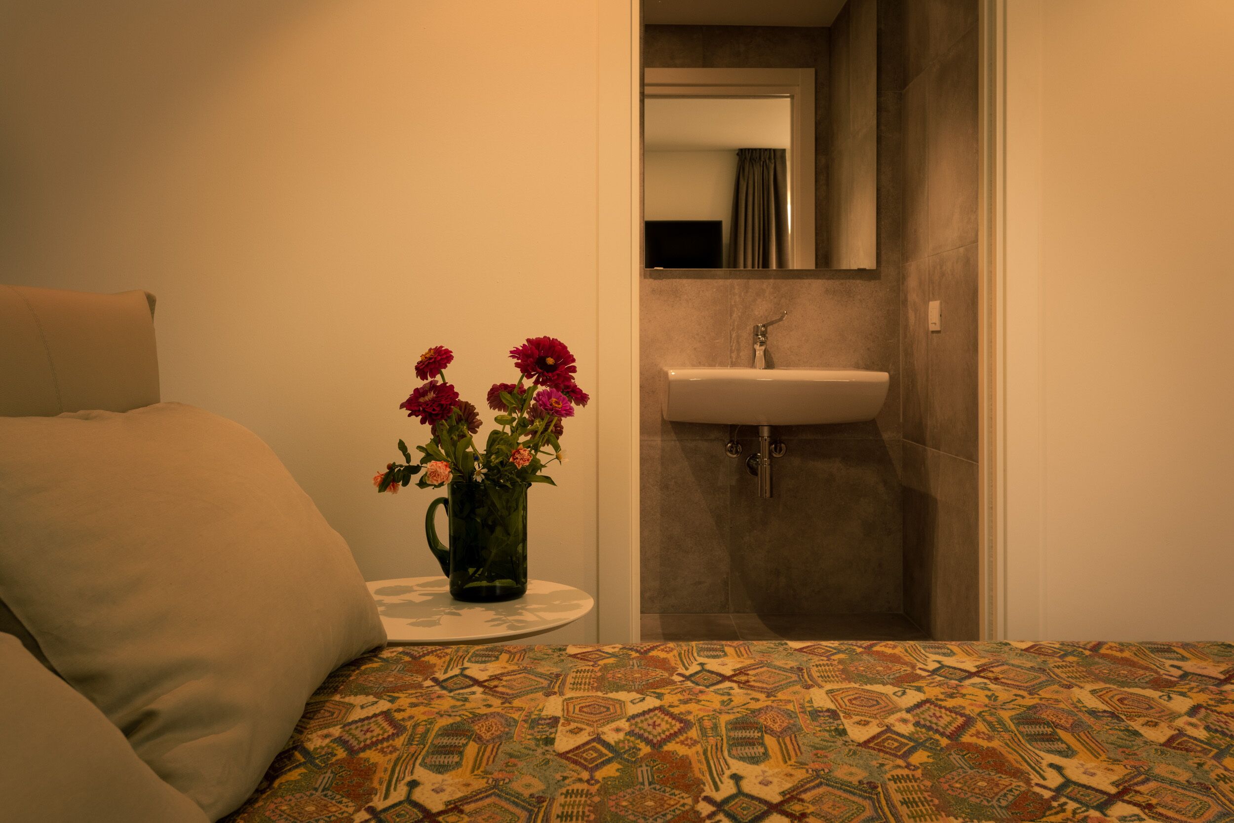 Quarto com cama, uma mesinha redonda com um vaso de flores e banheiro com pia e espelho.