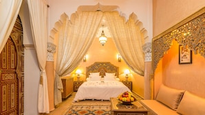 Um quarto luxuoso com uma cama ornamentada, um painel decorativo e uma fruteira sobre uma mesinha de cabeceira.
