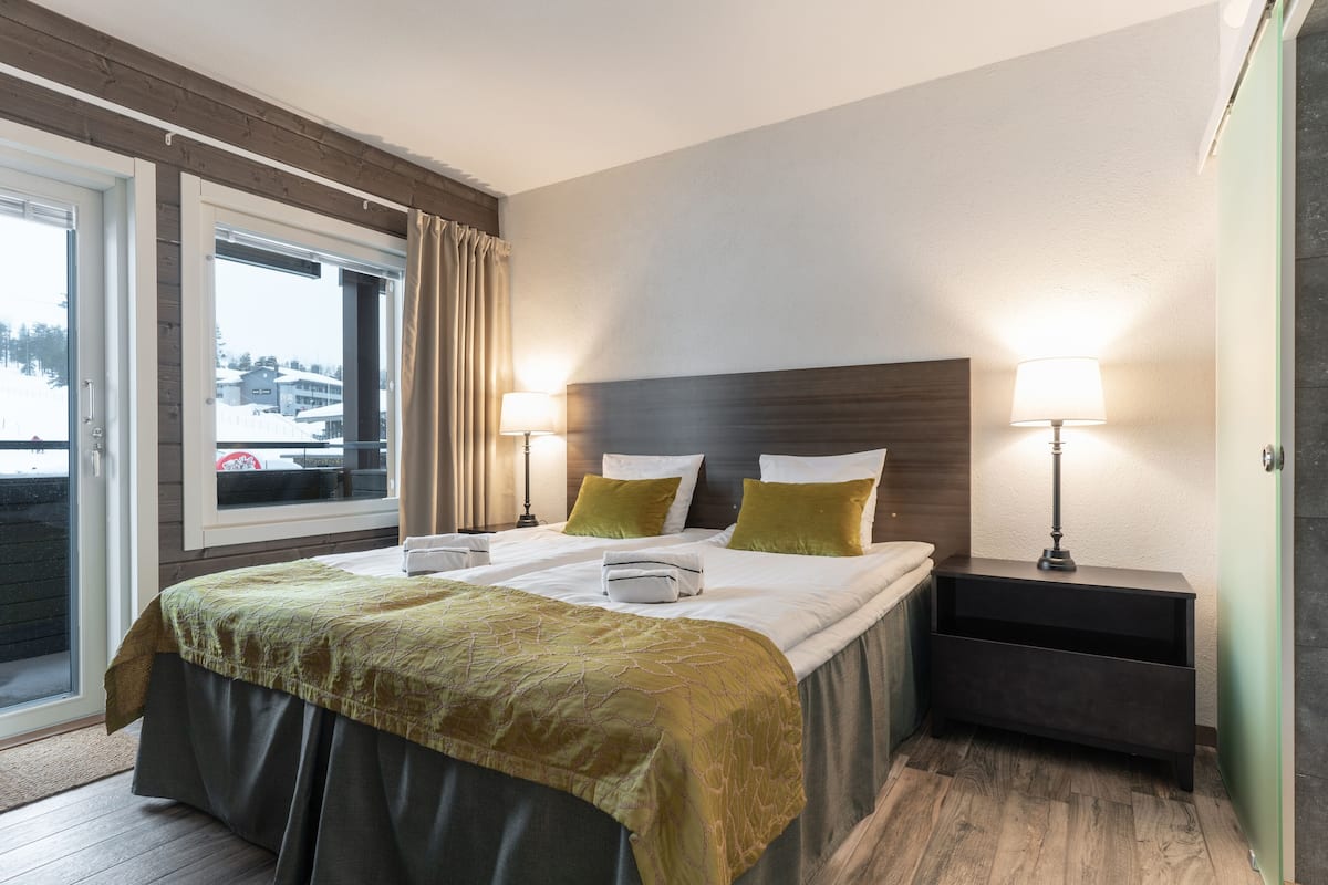 Quarto de hotel com uma cama grande, duas luminárias de cabeceira, um criado-mudo e uma janela com cortinas.