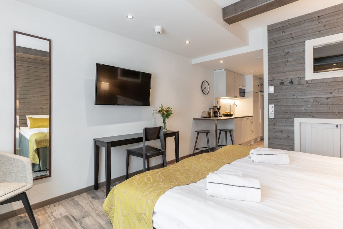 Quarto de hotel com cama, escrivaninha, cadeira, televisão e cozinha compacta.