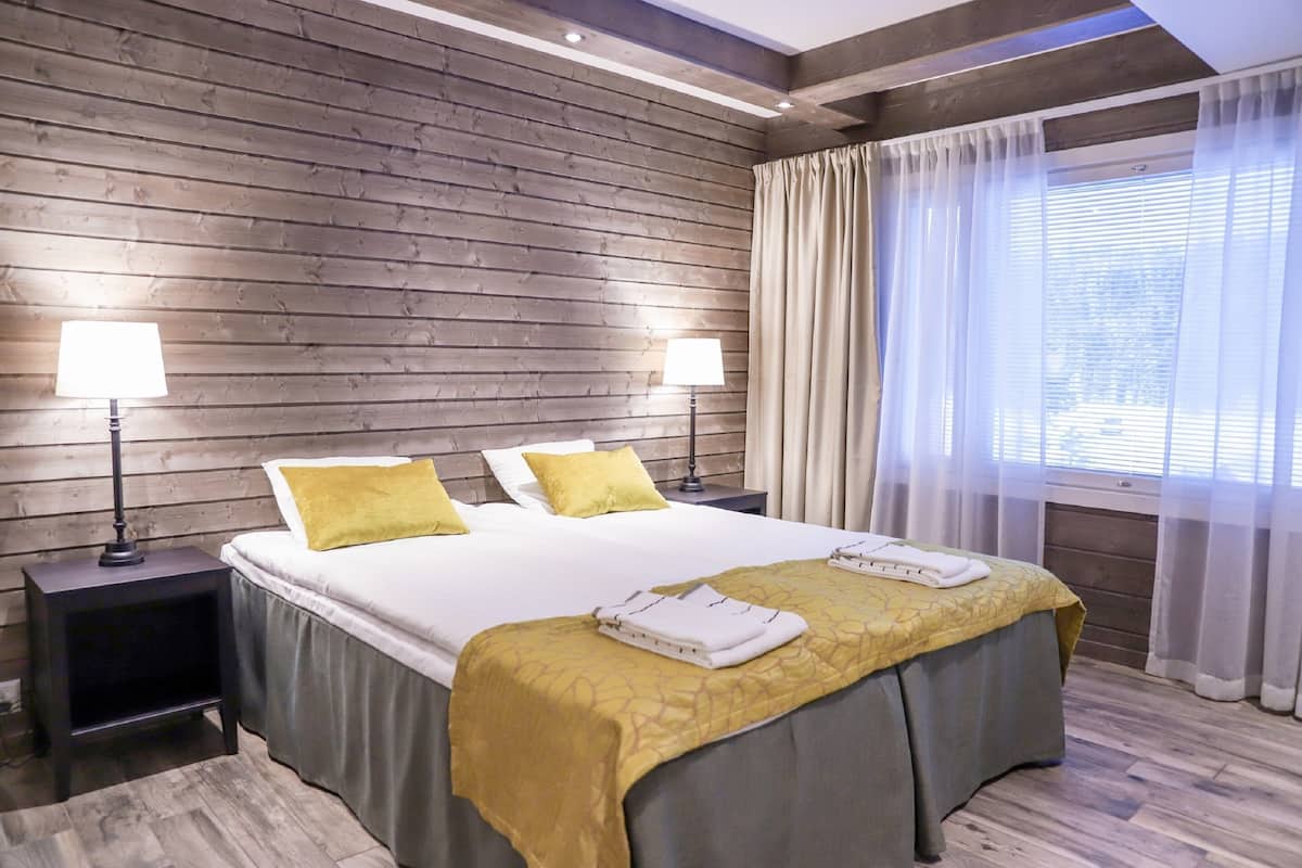 Quarto de hotel com parede de madeira, uma cama com colcha amarela, duas luminárias de mesa e uma janela com cortinas leves.