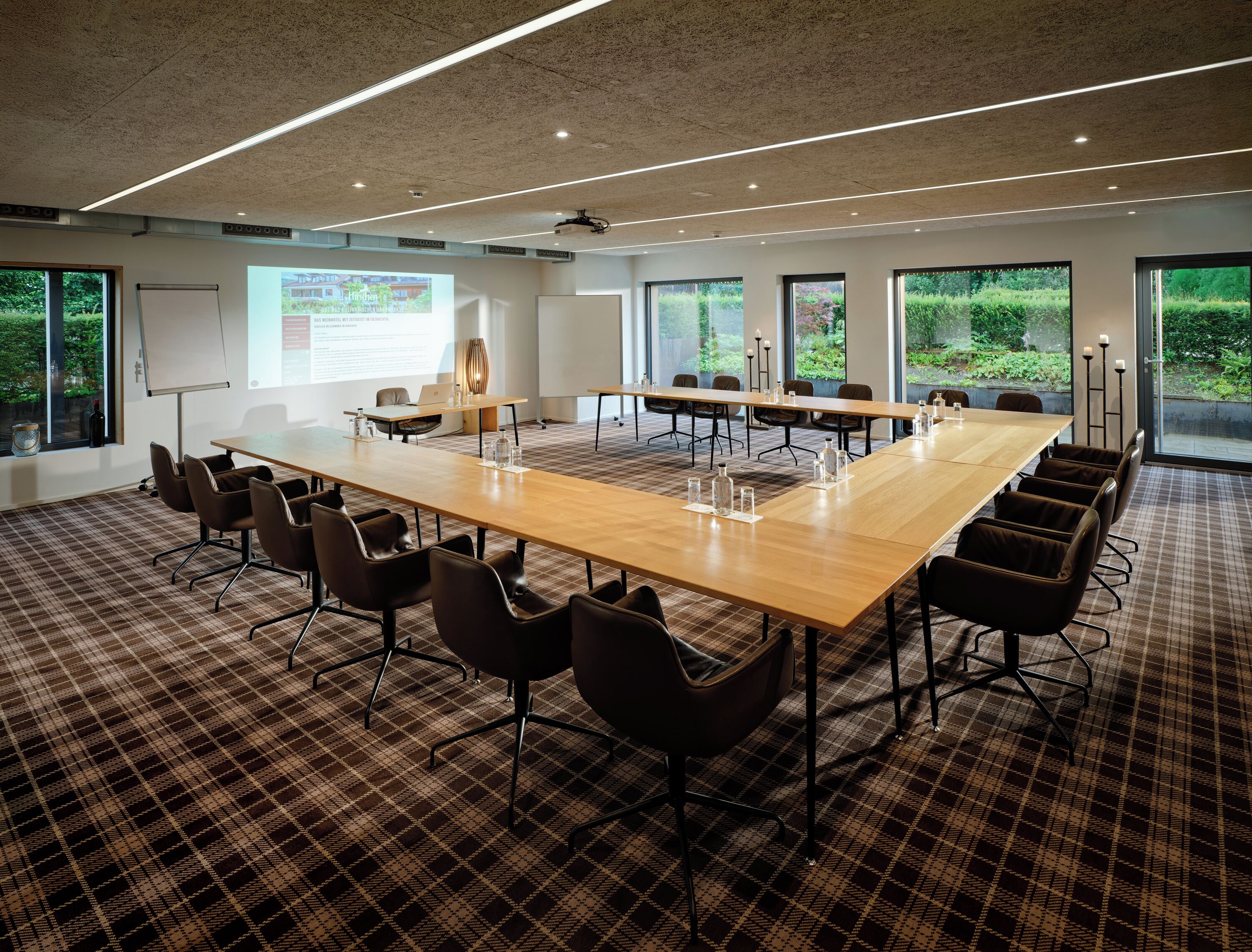 Sala de conferências com uma mesa comprida, cadeiras, tela de projetor e janelas amplas com vista para áreas verdes.