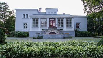 Um edifício grande e branco, de dois andares, com fachada simétrica, entrada central com porta vermelha e um jardim bem cuidado em primeiro plano.