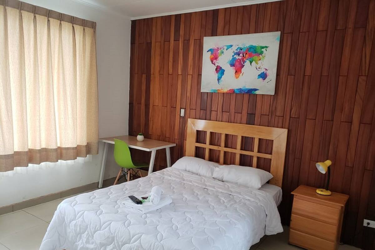 Quarto de hotel bem arrumado com cabeceira de madeira, cama com roupa de cama branca, pequena mesa de trabalho com cadeira, abajur e mapa-múndi na parede.