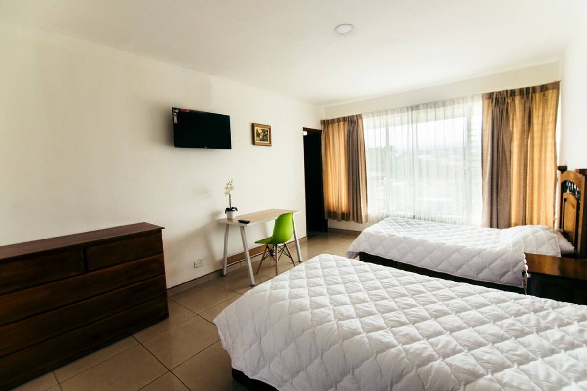 Quarto de hotel com duas camas, uma escrivaninha, uma cadeira, uma televisão fixada na parede e uma janela com cortinas.