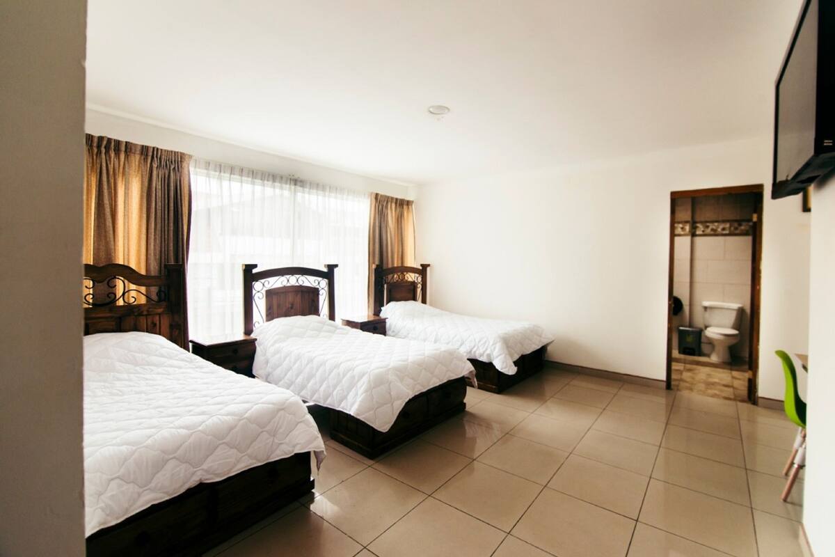 Quarto de hotel com duas camas de solteiro, cabeceira de madeira, uma mesinha, televisão fixada na parede e banheiro visível através de uma porta.