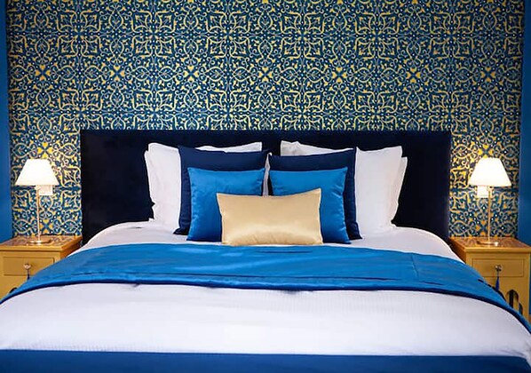 Cama com roupa de cama azul e branca, parede com padrão azul e amarelo, e duas luminárias de cabeceira.
