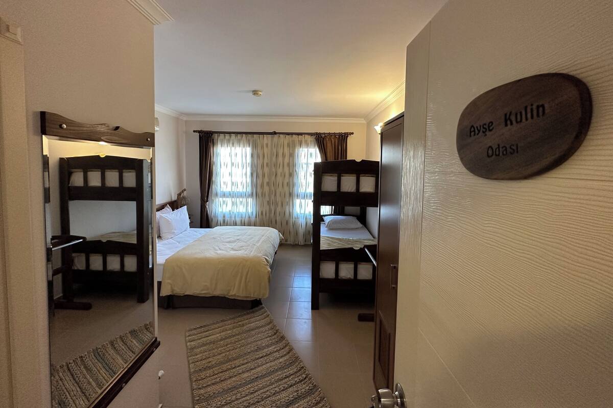 Quarto de hotel com uma cama grande, duas camas menores, cabeceira de madeira, uma placa de identificação e vista para o exterior.