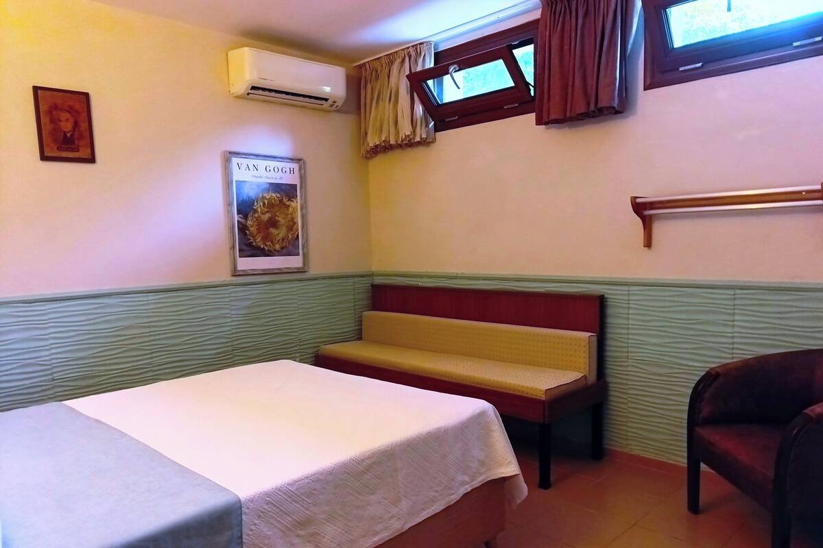 Um quarto com cama, um banco, uma cadeira, uma janela com cortinas, um aparelho de ar condicionado e um cartaz na parede.