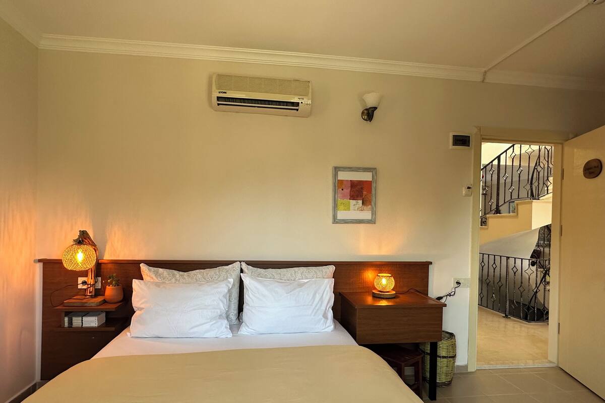 Um quarto com uma cama, duas mesas de cabeceira, um ar condicionado fixado na parede, um quadro emoldurado e uma escada que leva a um andar superior.