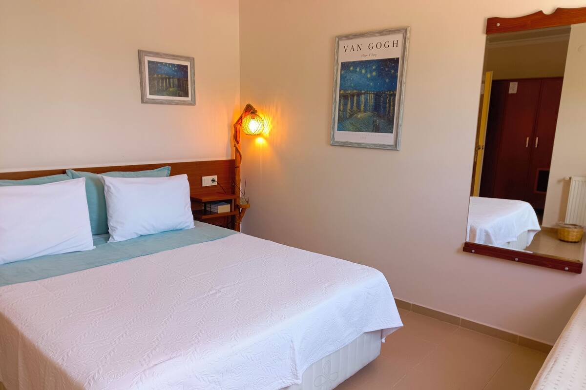 Quarto de hotel com uma cama grande, uma mesa de cabeceira com luminária, uma pintura de Van Gogh emoldurada e um espelho refletindo o quarto.