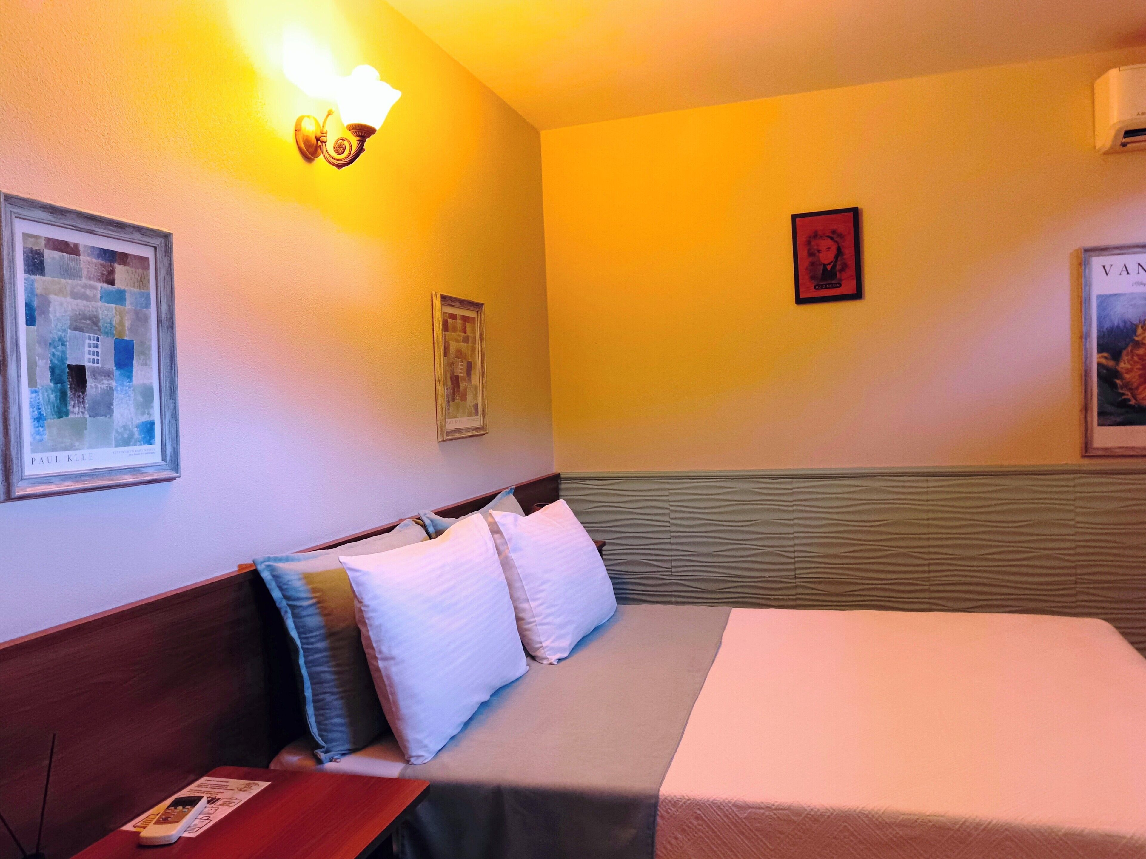 Quarto de hotel com cama, mesa de cabeceira, abajur e obras de arte emolduradas nas paredes.