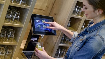 Uma pessoa interagindo com um menu digital em um bar de sucos, com prateleiras de recipientes de vidro ao fundo.