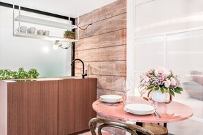 Uma sala de jantar com uma mesa redonda, cadeiras e toalha de mesa rosa. Há um armário de madeira com uma prateleira que acomoda xícaras e uma planta. Um vaso com flores está sobre a mesa.