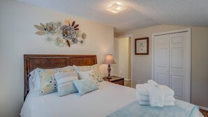 Cama bem arrumada com almofadas florais, luminária de cabeceira e quadro na parede.