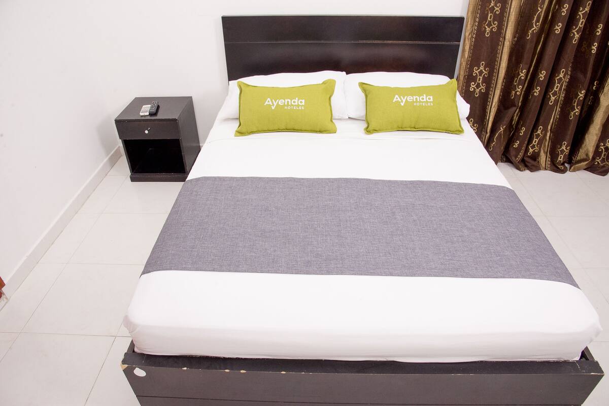 Cama bem arrumada com um cobertor cinzento e duas almofadas verdes com a inscrição "Ayenda Hotels", um criado-mudo preto com um controle remoto e uma cortina marrom com padrão.