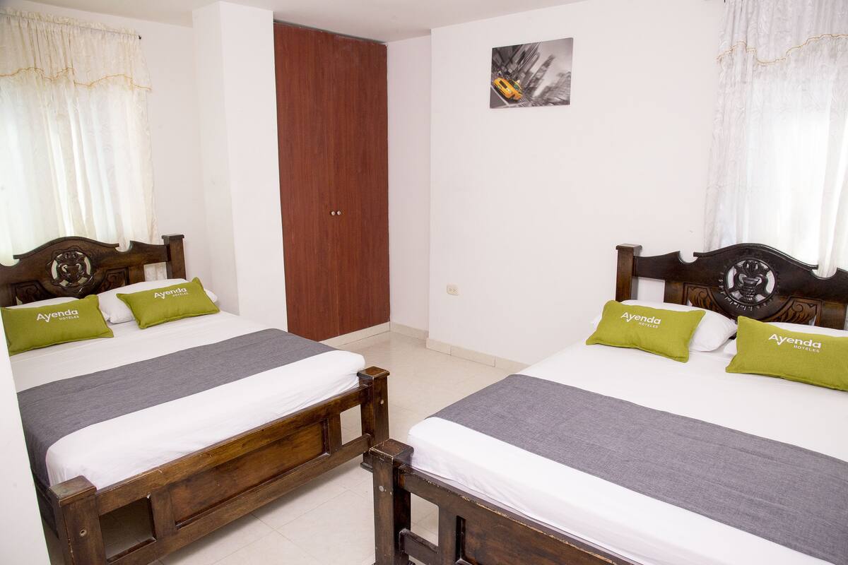 Quarto de hotel com duas camas de madeira, cada uma com colcha cinza e branca, e travesseiros verdes com o nome do hotel.