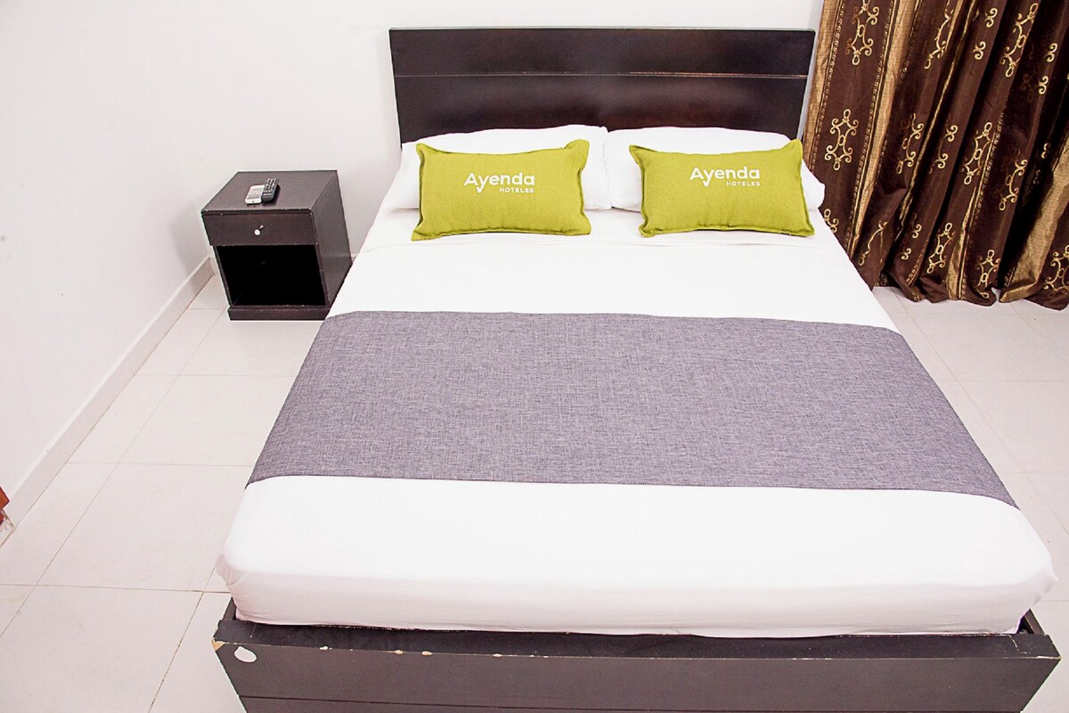 Uma cama bem arrumada com duas almofadas, uma mesa de cabeceira com um controle remoto e uma cabeceira escura.