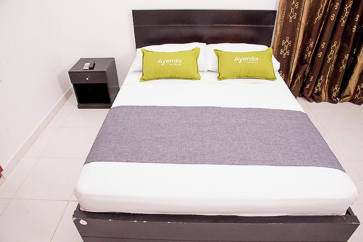 Uma cama bem arrumada com duas almofadas, uma mesa de cabeceira com um controle remoto e uma cabeceira escura.
