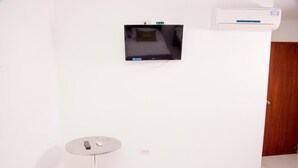 Quarto com televisão de tela plana na parede, uma mesa redonda pequena com um controle remoto e um aparelho de ar condicionado acima da TV.