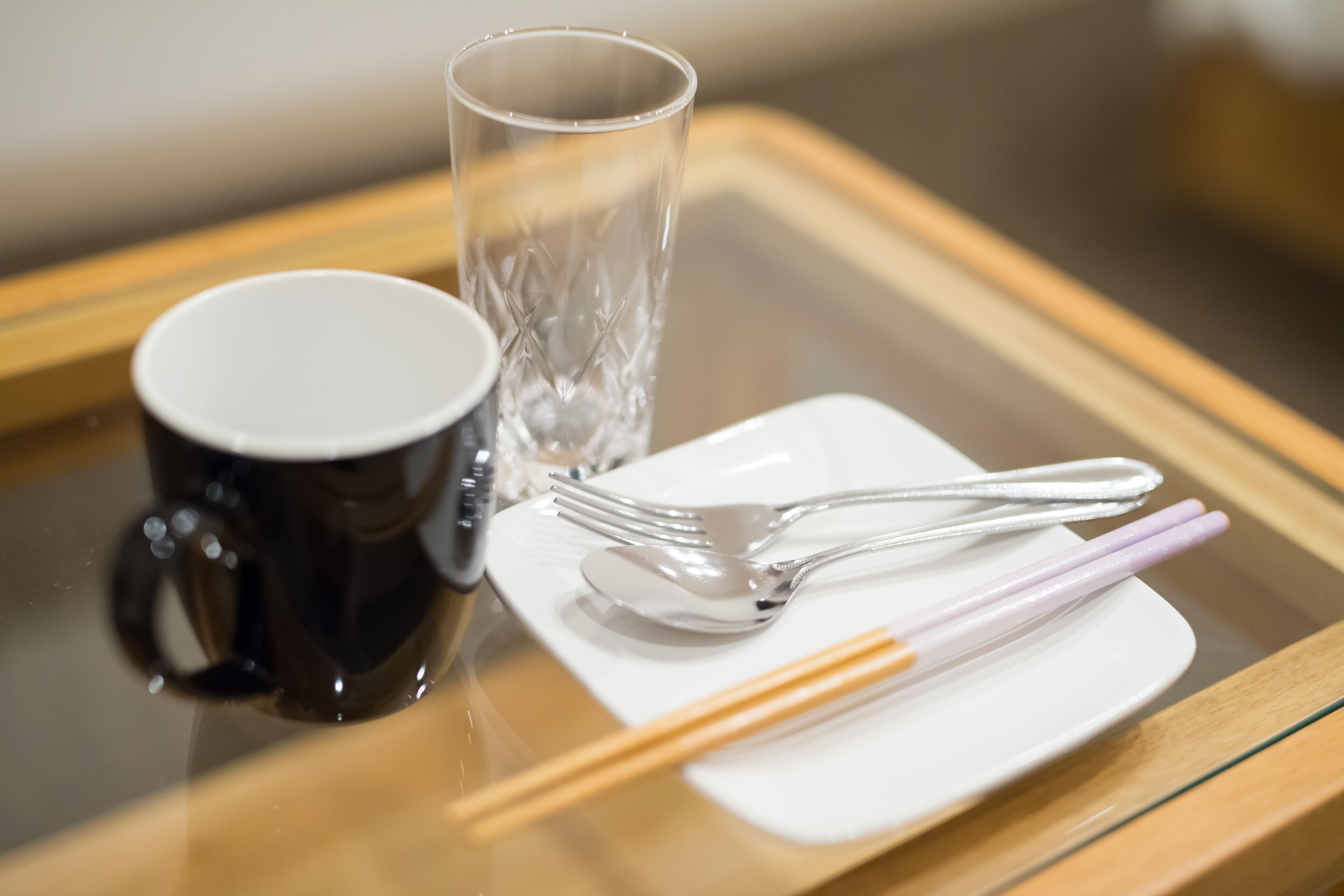 Uma bandeja com uma caneca preta, um copo transparente, um prato branco com uma colher e hashis, e um garfo.
