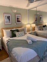 Uma cama bem arrumada com um cobertor verde, uma toalha branca e uma cabeceira de madeira. Há duas mesas de cabeceira com luminárias e um espelho grande na parede.