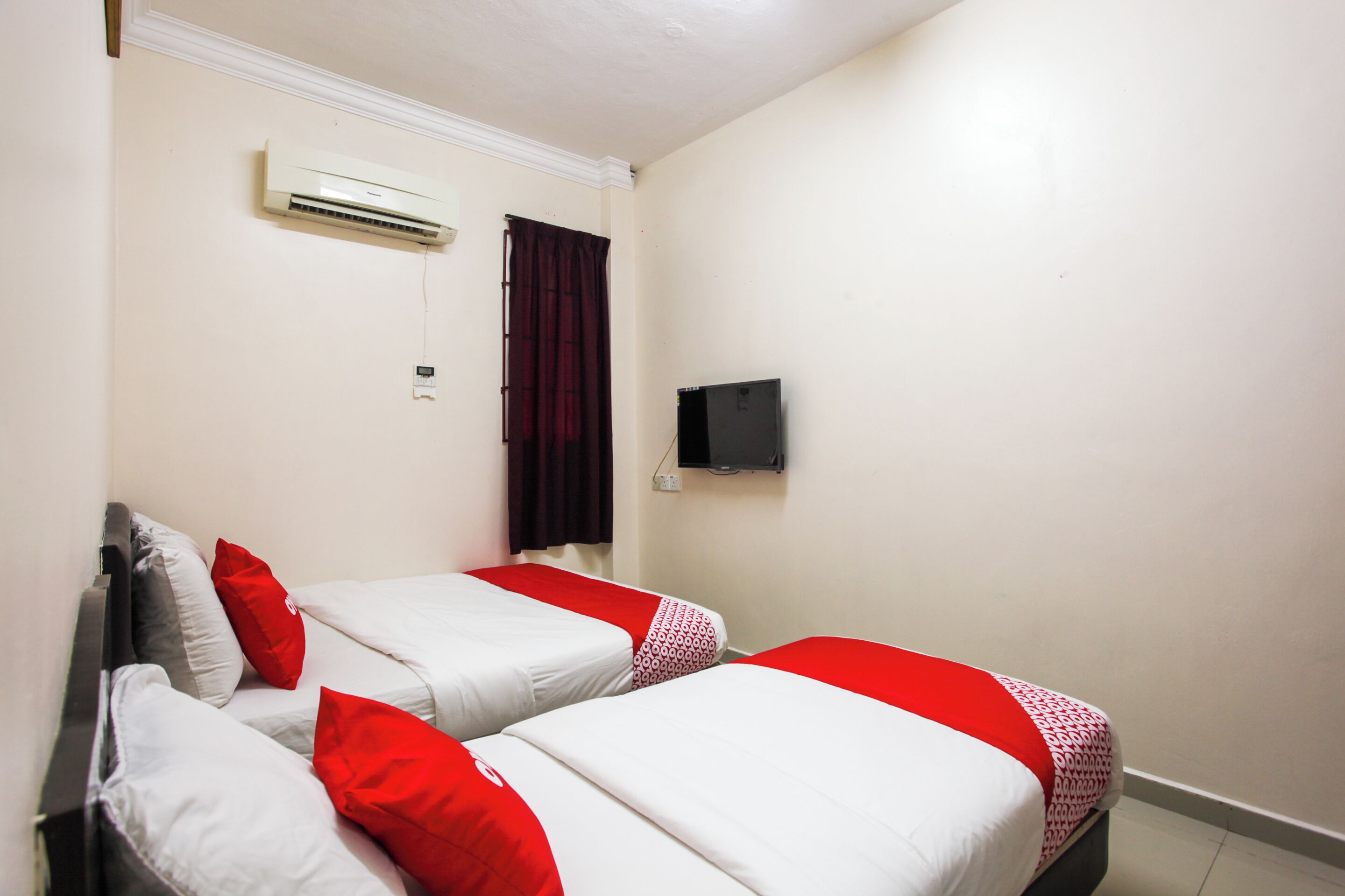 Quarto com duas camas, cada uma com roupa de cama vermelha e branca, uma televisão na parede e um ar condicionado.