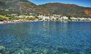 Uma vila costeira com águas azul-claras, edifícios brancos e montanhas ao fundo.