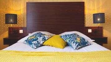 Cama bem arrumada com um cobertor amarelo, duas almofadas florais e uma cabeceira de madeira. Há duas mesas de cabeceira com abajures e uma parede com um padrão decorativo.